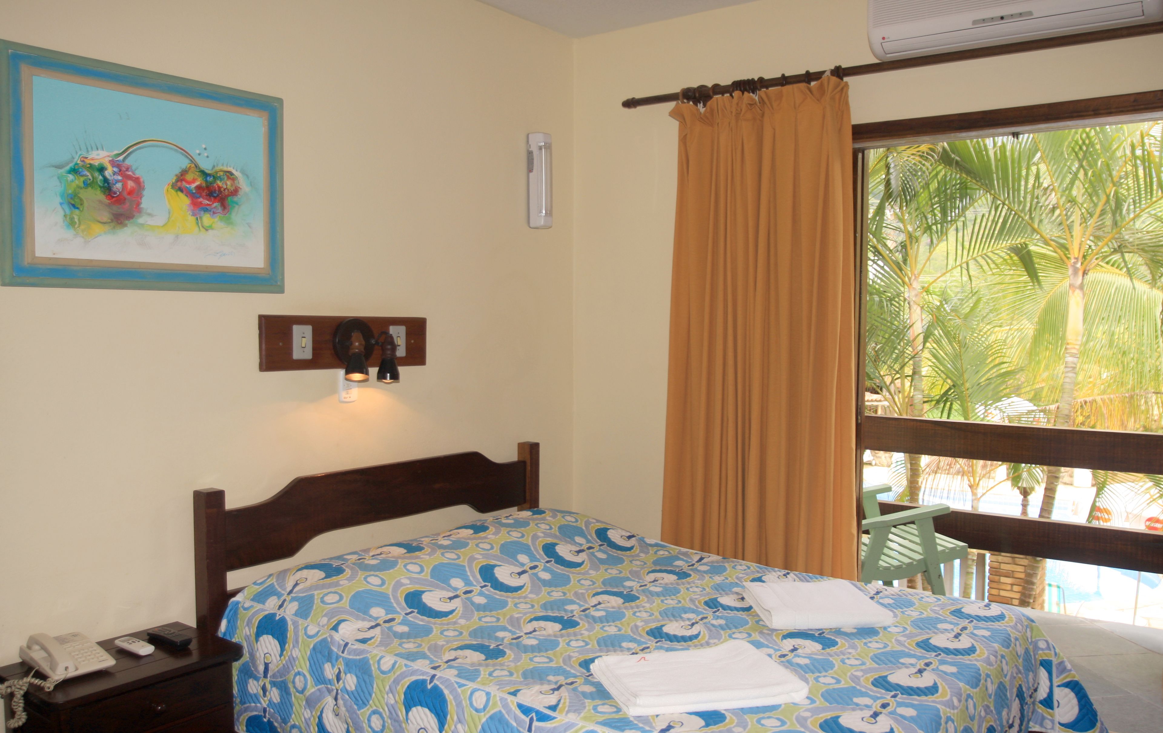 Quarto de hotel com uma cama, mesa de cabeceira com telefone e controle remoto, quadro na parede, janela com cortinas e vista para palmeiras.