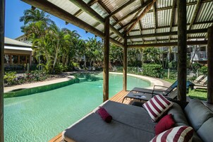Uma área à beira da piscina com um espaço de estar coberto, piscina e vegetação tropical ao redor.