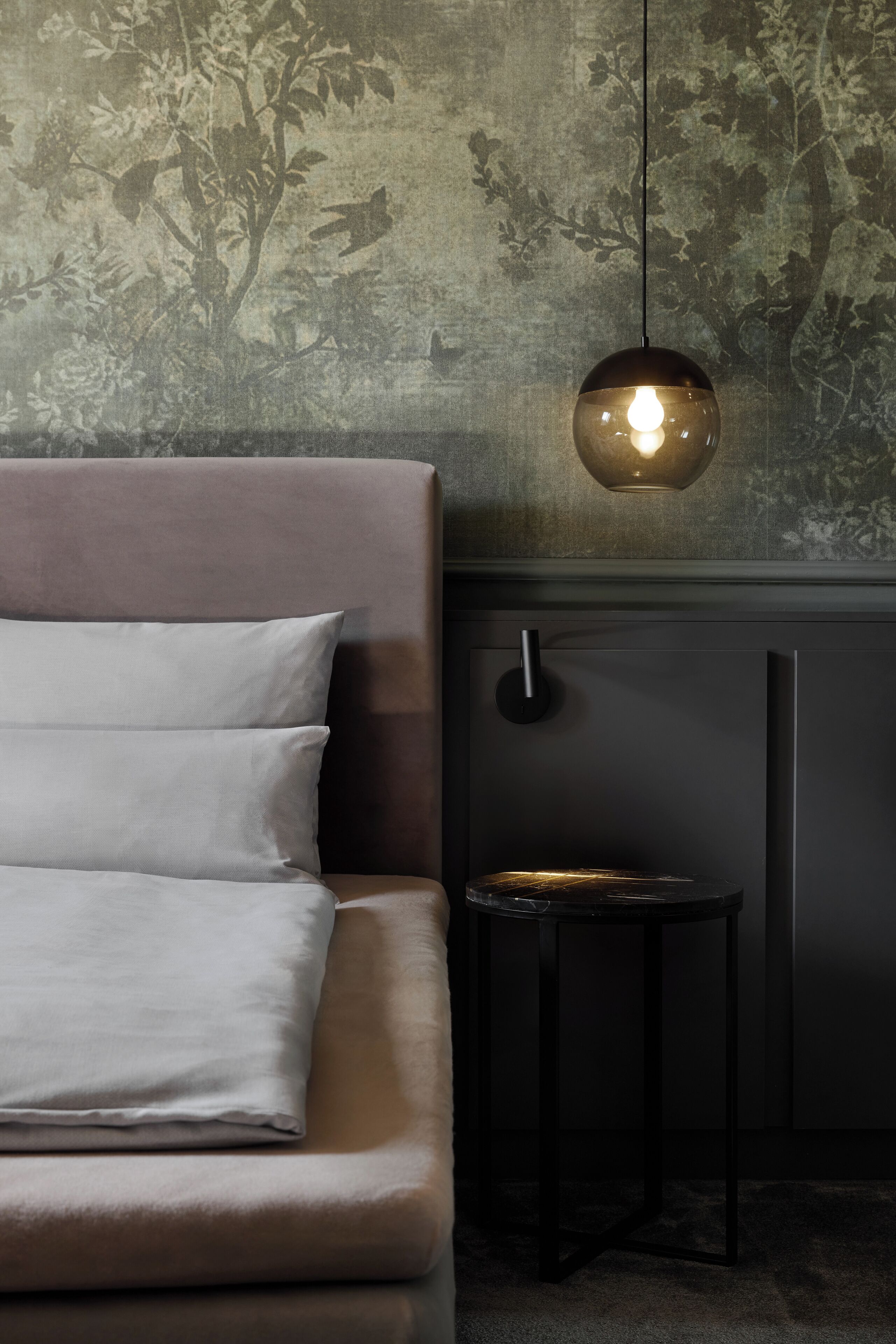 Quarto com cama, uma mesinha e luminária fixada na parede.