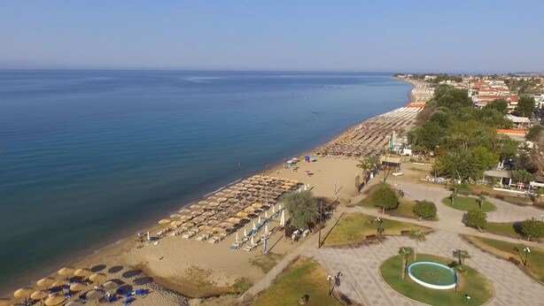 Vista aérea de uma praia com fileiras de guarda-sóis e cadeiras de praia, um calçadão com uma fonte e uma área residencial ao fundo.