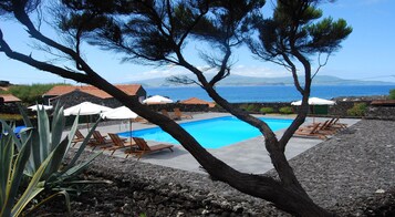 Área de piscina com espreguiçadeiras e guarda-sóis, cercada por terreno rochoso e pinheiros, com vista para a paisagem costeira.