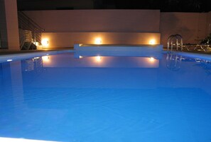 Piscina com superfície de água azul cristalina, escada de metal e parede iluminada ao fundo.