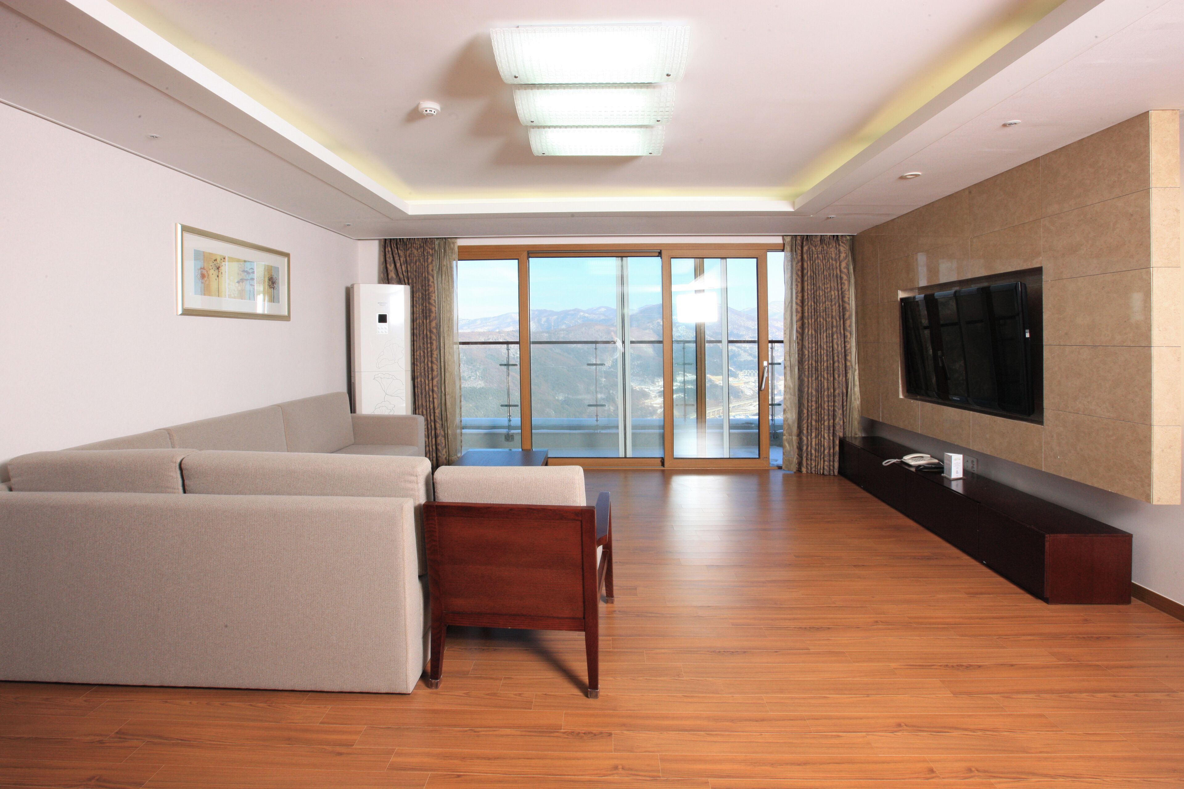 Uma sala de estar moderna com sofá, mesa de centro de madeira, TV de tela plana na parede e uma grande janela com vista para as montanhas.