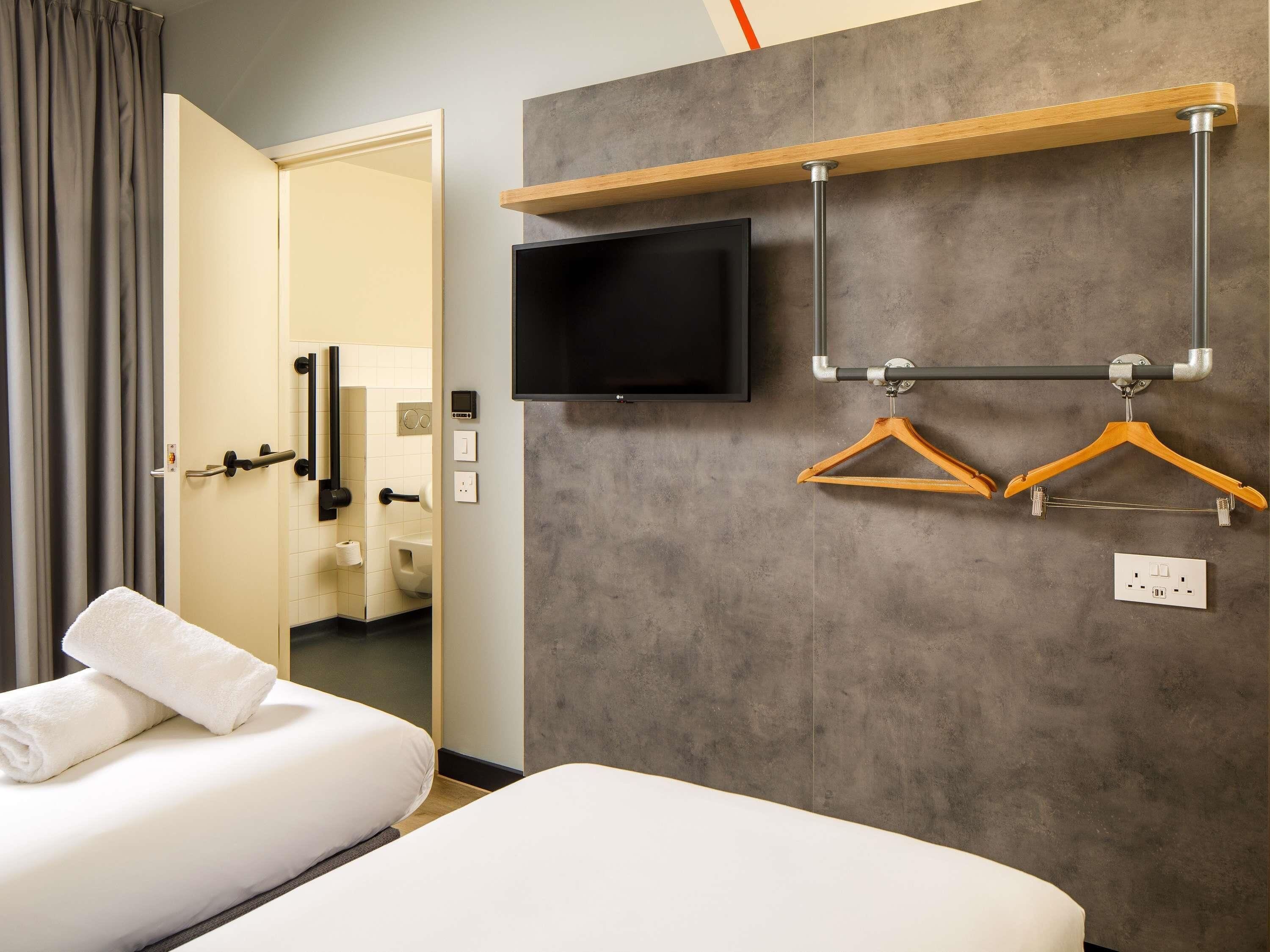Quarto de hotel com cama, televisão na parede, prateleira de madeira e toalha sobre a cama.