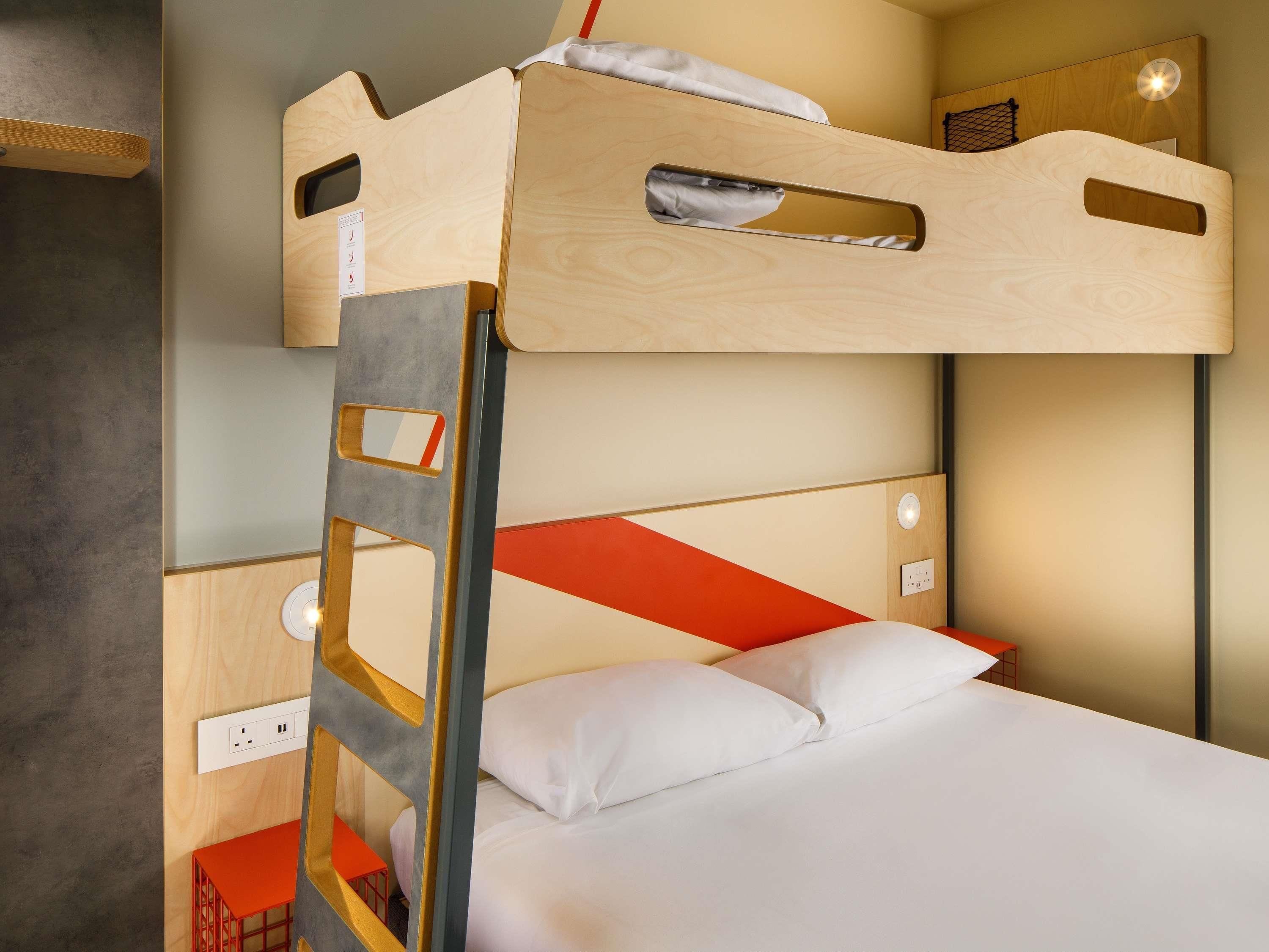 Uma cama de beliche com escada, uma mesinha de cabeceira e uma tomada na parede.