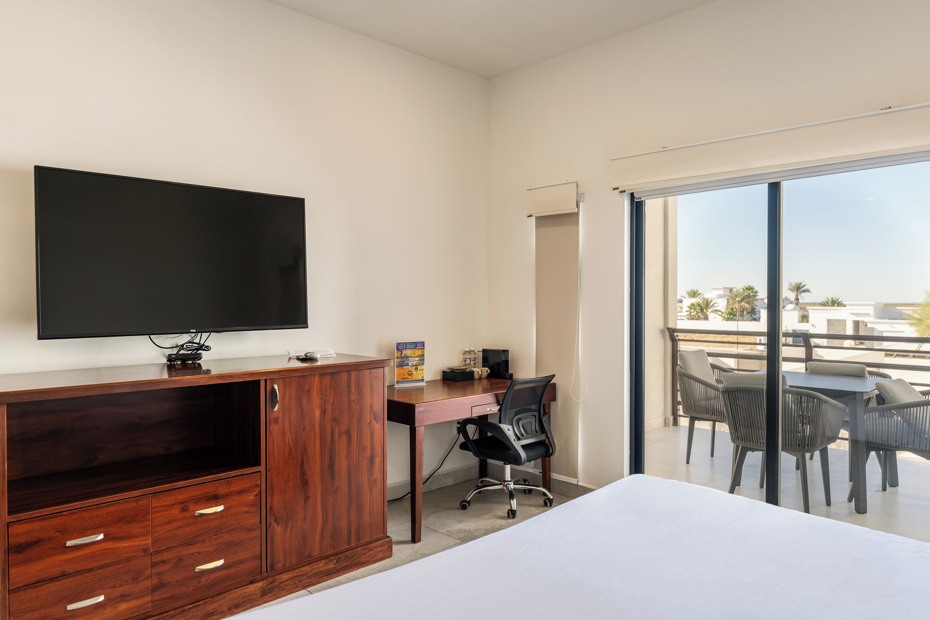 Quarto de hotel com televisão grande, móveis de madeira, secretária com cadeira e varanda com área de estar externa.