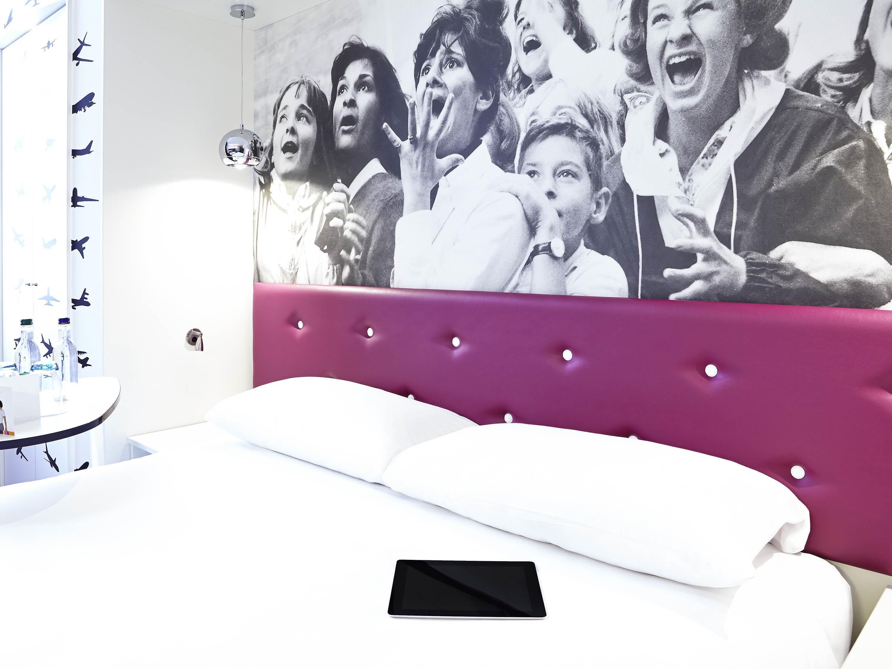 Quarto com cama, mesa de cabeceira com garrafas e parede com uma foto grande.