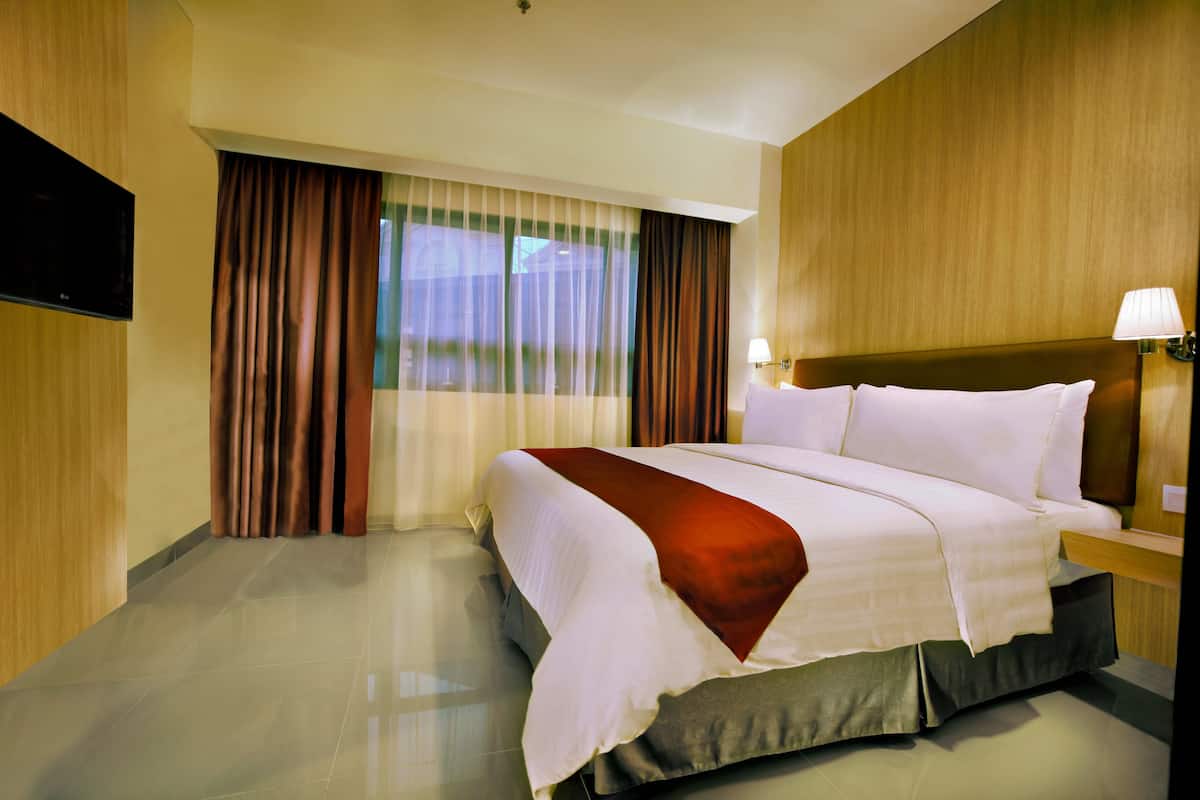 Quarto de hotel com cama grande, televisão de tela plana na parede, mesa de cabeceira com luminária e janela com cortinas.