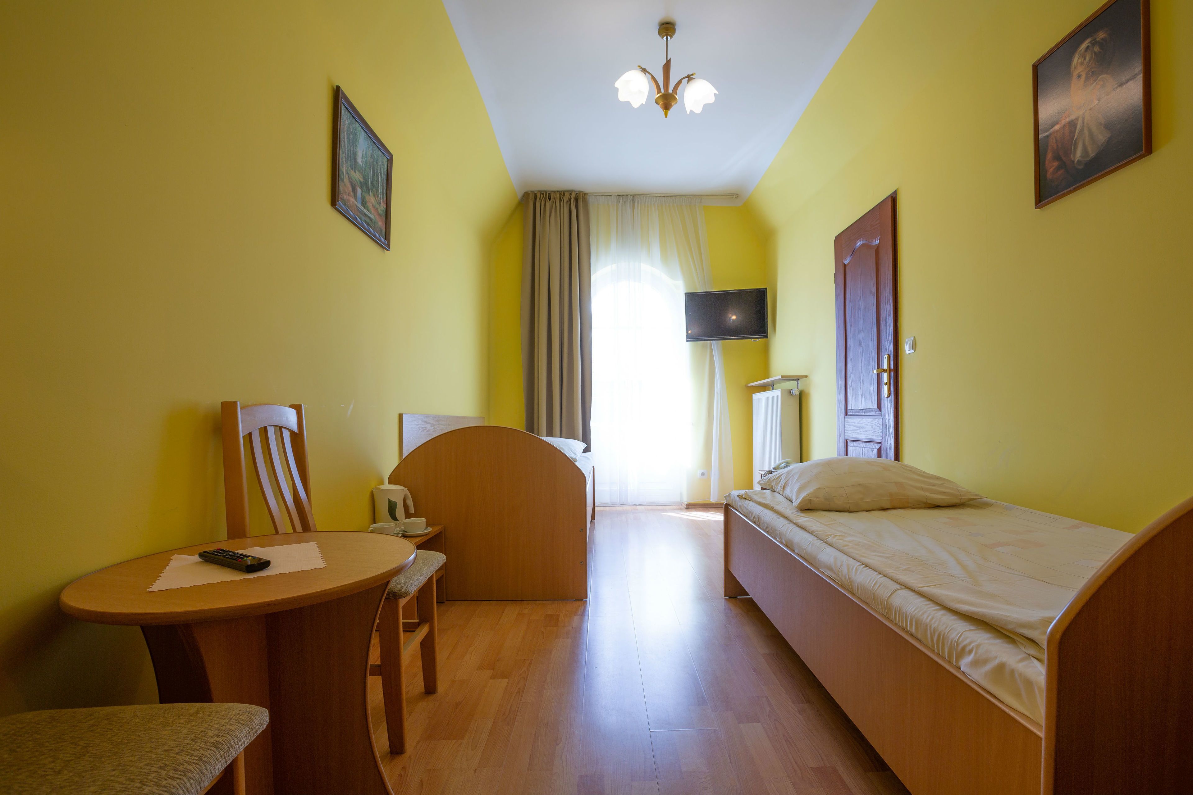 Quarto de hotel pequeno com uma cama de solteiro, uma mesa com uma cadeira, televisão e um quadro na parede.