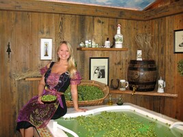 Mulher vestida com traje tradicional segura uma cesta de folhas verdes, posicionada ao lado de uma banheira cheia de folhas verdes, com paredes e prateleiras de madeira exibindo diversos frascos e um barril grande.