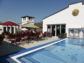 Uma área de piscina com espreguiçadeiras e guarda-sóis, um edifício branco com emblema dourado e uma pequena estrutura em torre.