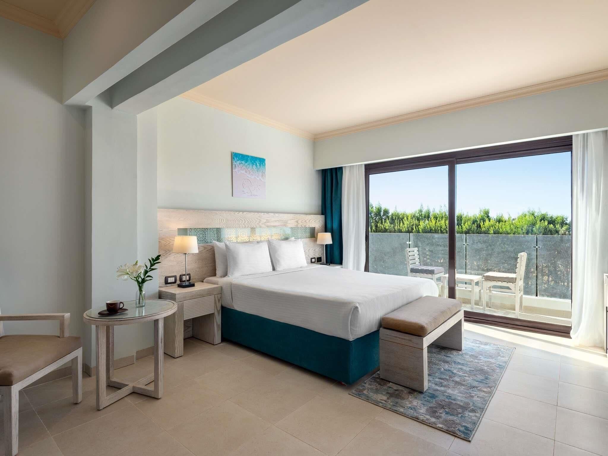 Quarto de hotel com uma cama grande, uma mesinha com uma luminária, uma cadeira e vista para o verde através de uma varanda.