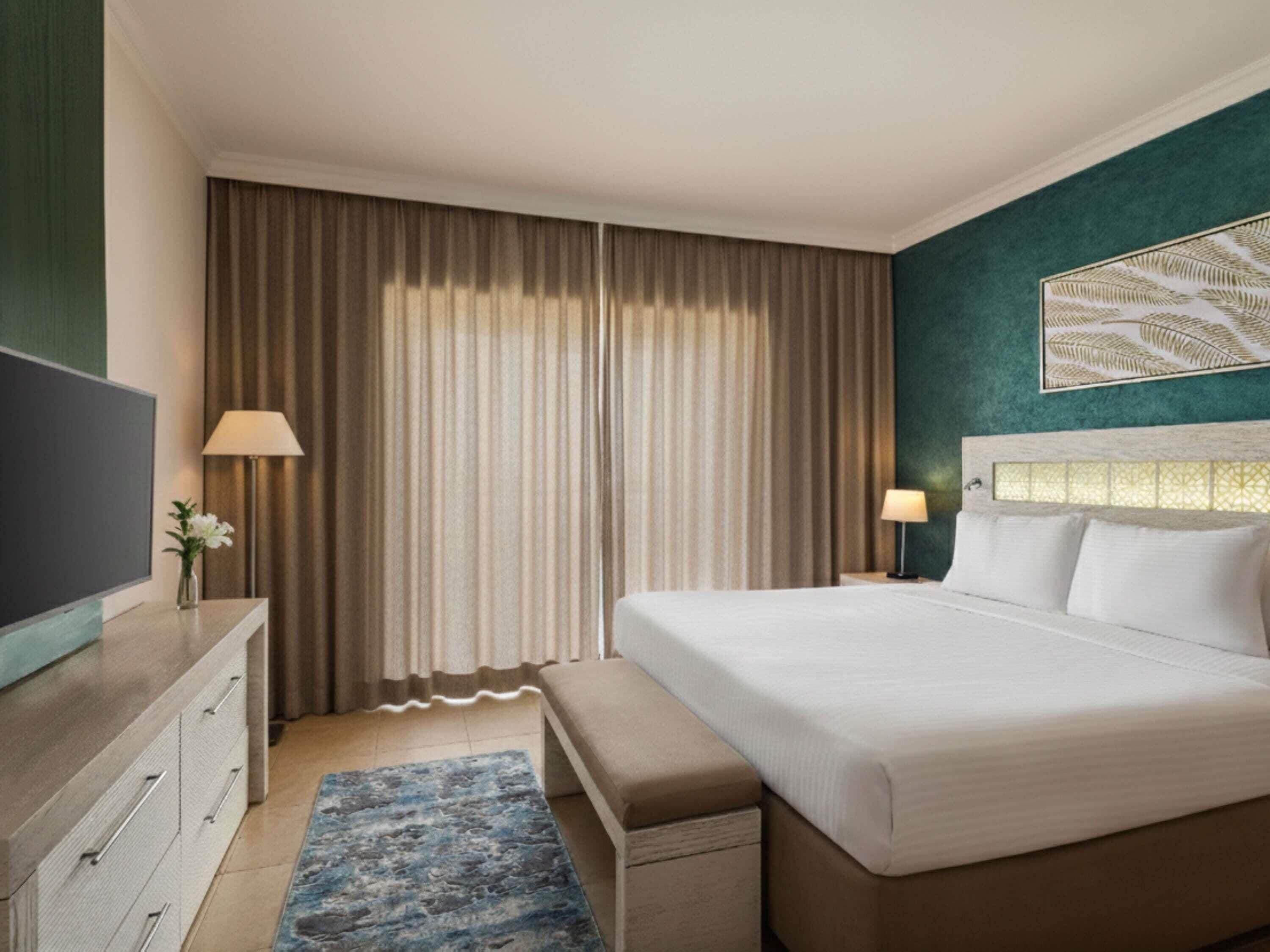 Quarto de hotel com uma cama grande, televisão de tela plana, mesa de cabeceira com abajur e um objeto decorativo na parede.