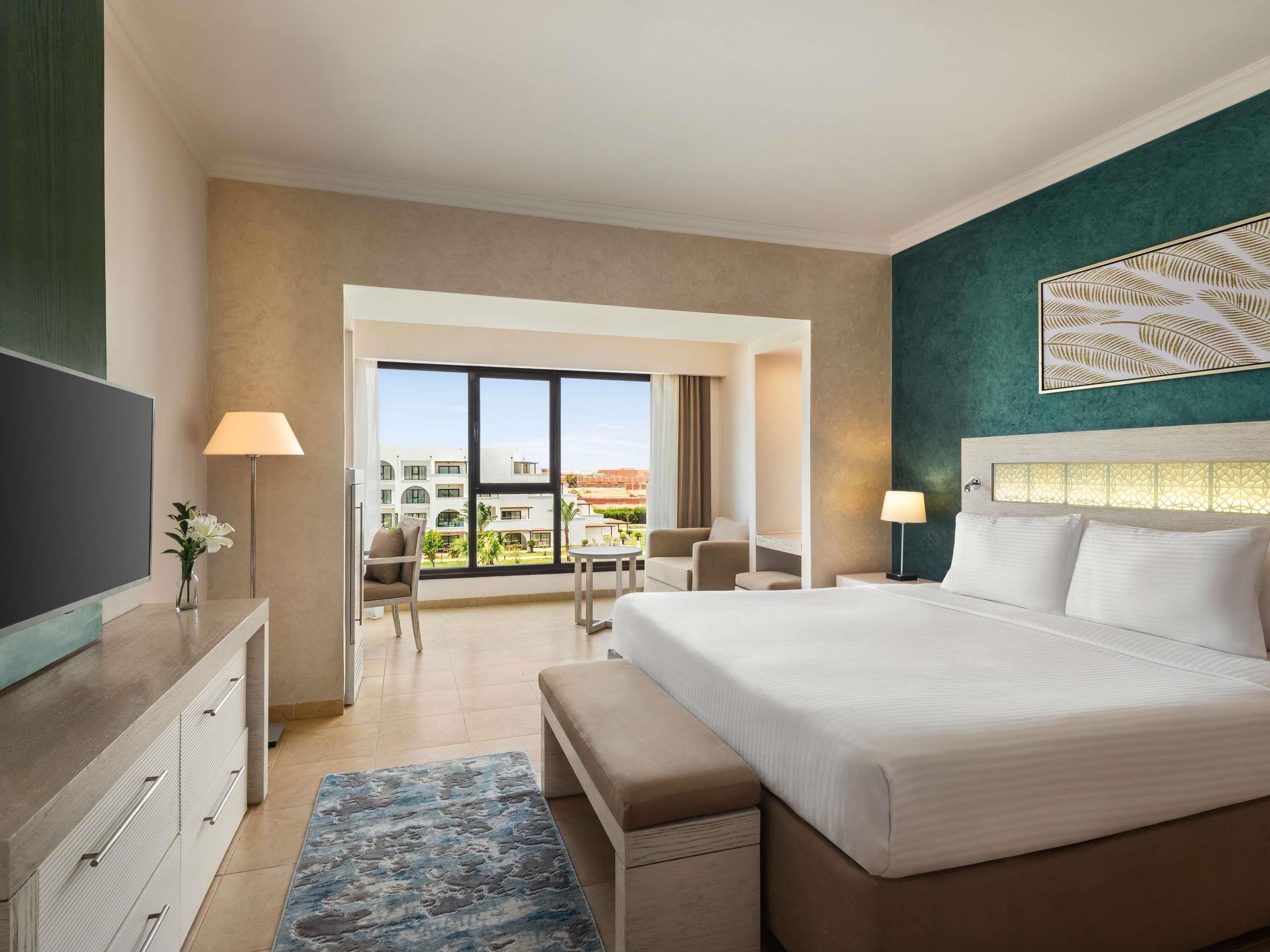 Quarto de hotel com cama, mesas de cabeceira, televisão, escrivaninha, cadeira, abajur e vista para edifícios através de grandes janelas.