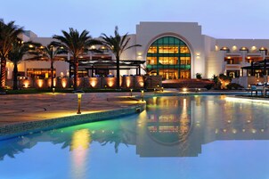 Um hotel com uma piscina grande, palmeiras e uma entrada bem iluminada.