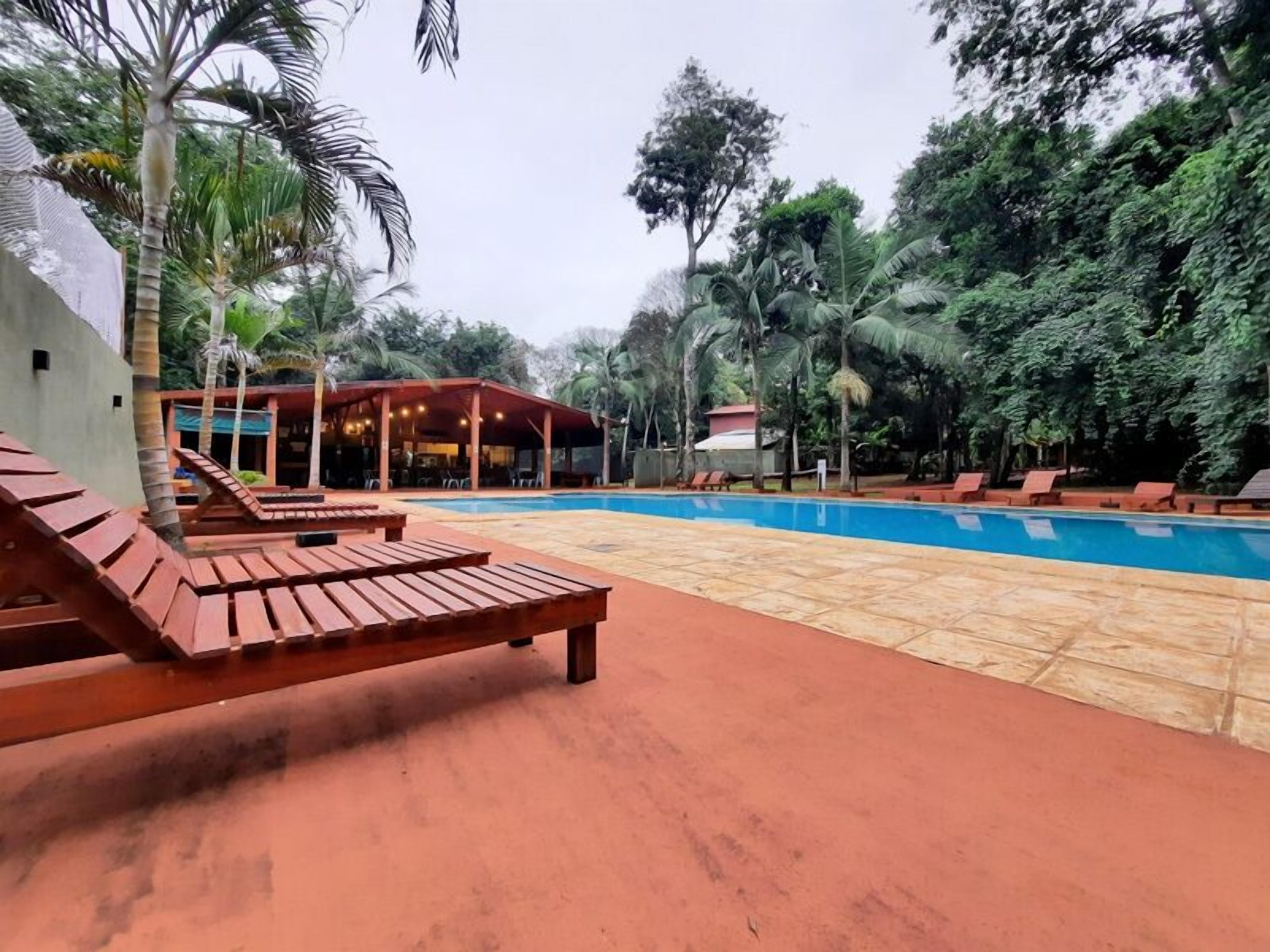 Uma área de piscina com espreguiçadeiras de madeira, um espaço de estar coberto e vegetação exuberante.