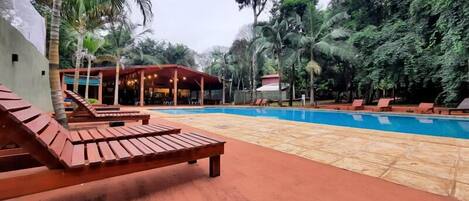Uma área de piscina com espreguiçadeiras de madeira, um espaço de estar coberto e vegetação exuberante.
