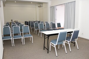 Uma sala de conferências com fileiras de cadeiras e uma mesa, piso coberto de carpete e uma janela com cortinas.