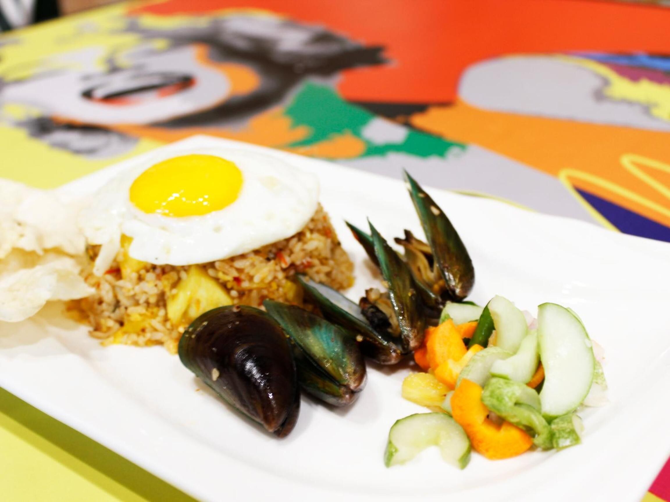 Um prato de arroz frito coberto com ovo frito, mariscos e uma guarnição de legumes fatiados.