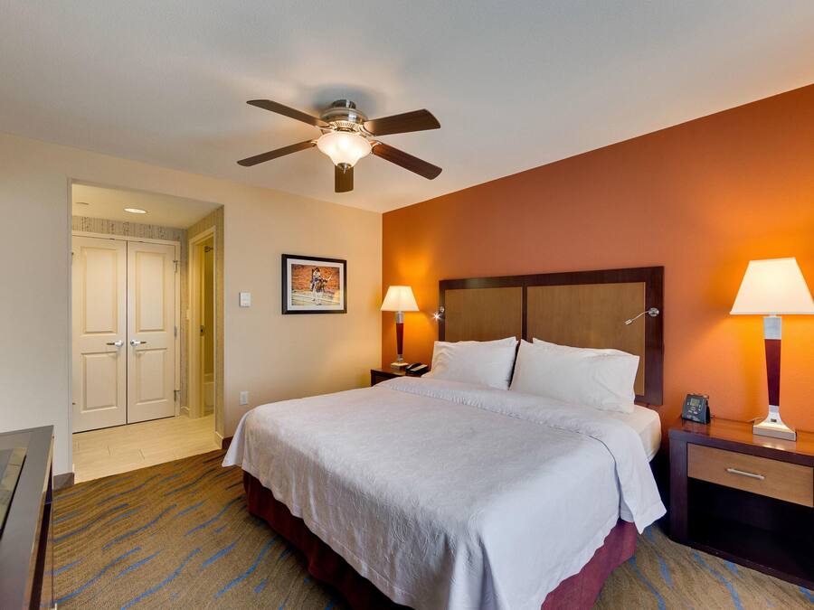 Quarto de hotel com cama, duas luminárias de cabeceira, mesa de cabeceira, ventilador de teto, porta e piso coberto por carpete.
