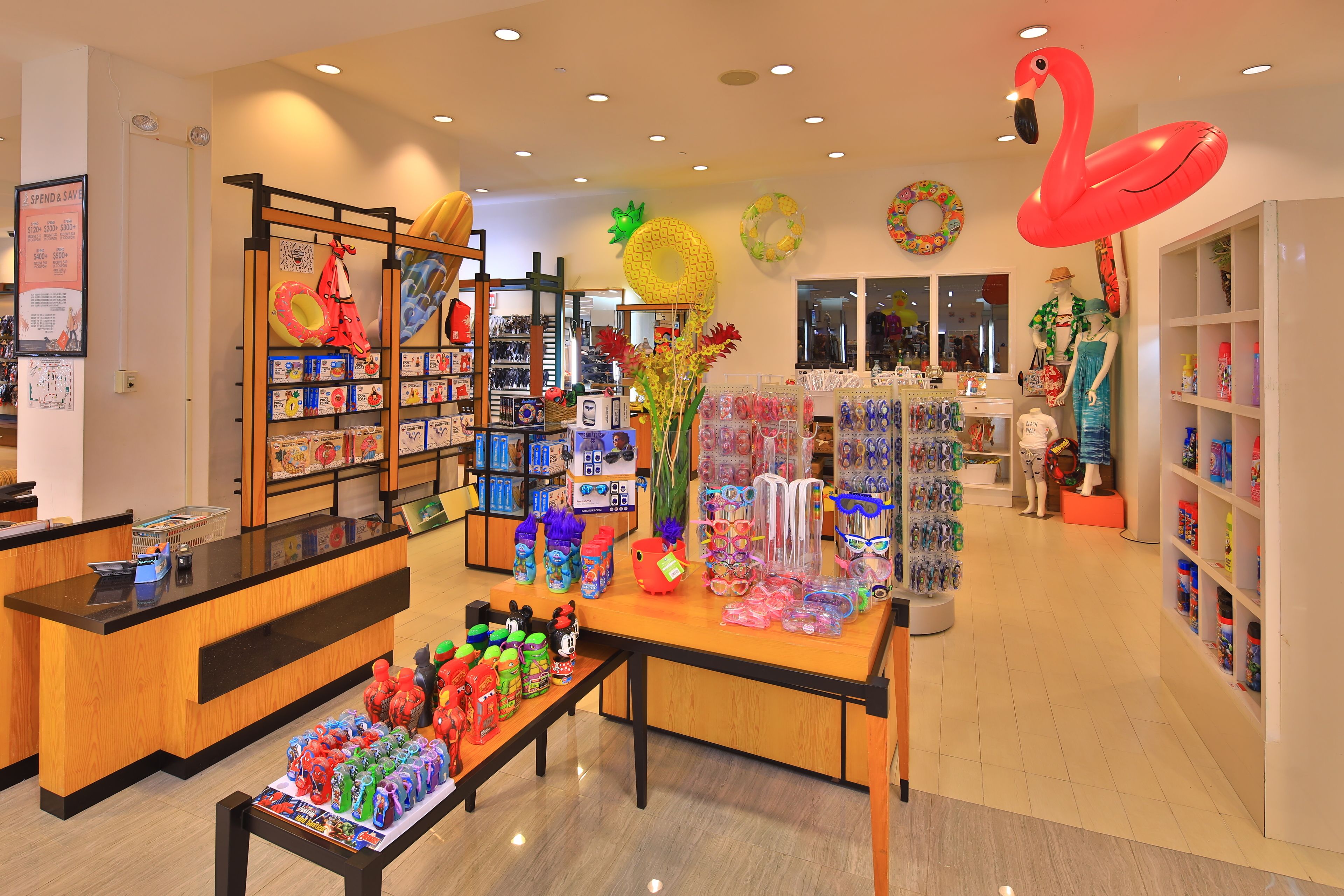 Loja bem iluminada com prateleiras cheias de brinquedos e acessórios coloridos, uma grande decoração de flamingo e uma vitrine com diversos itens, incluindo um enfeite em formato de abacaxi.