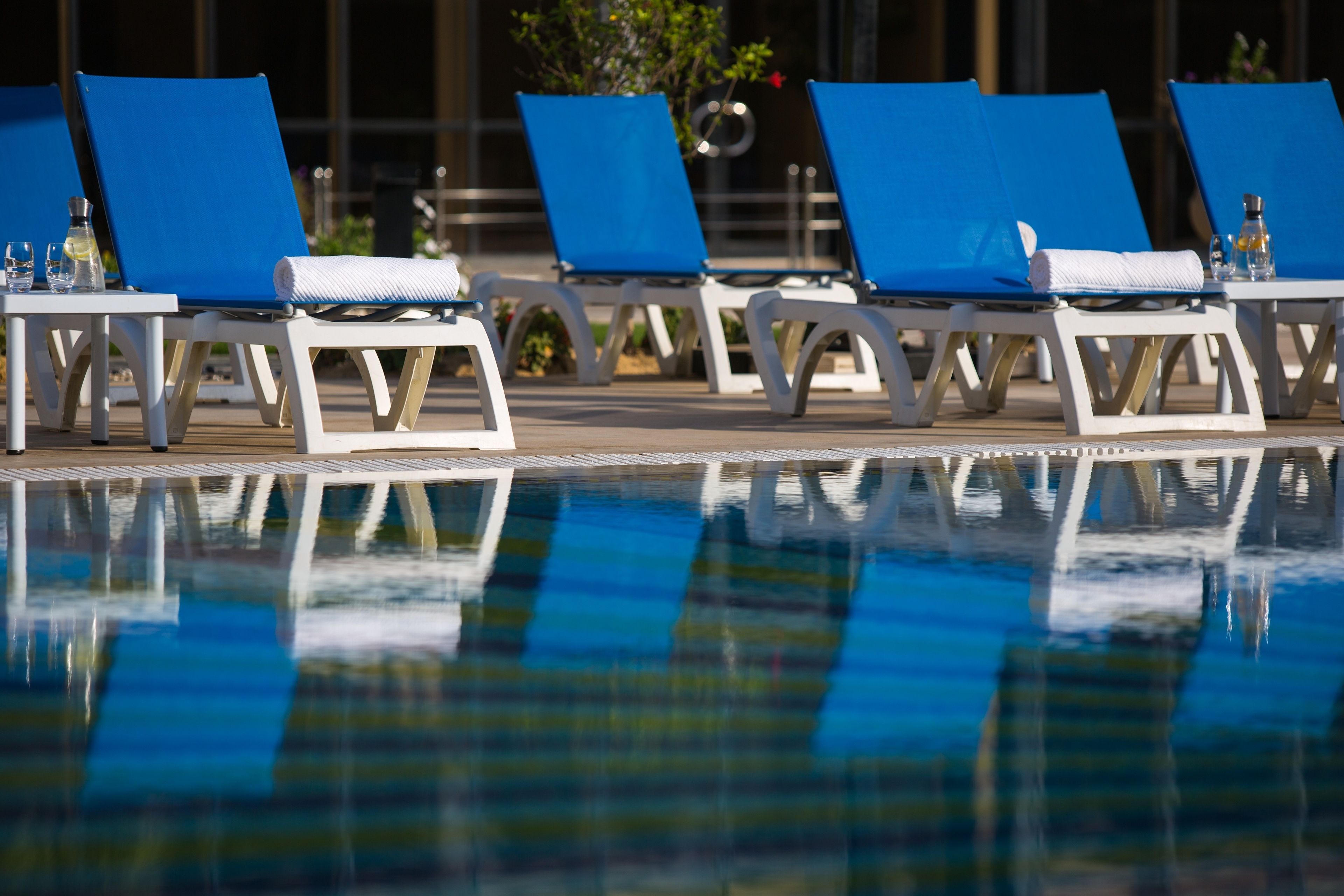Área da piscina com espreguiçadeiras e mesas, piscina cristalina e um edifício ao fundo.