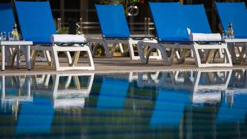 Área da piscina com espreguiçadeiras e mesas, piscina cristalina e um edifício ao fundo.