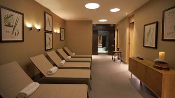 Uma sala de spa com três macas de massagem, uma mesa lateral de madeira com uma vela e impressões botânicas emolduradas nas paredes.