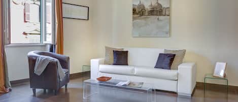 Uma sala de estar com um sofá branco, uma poltrona marrom, uma mesa de centro de vidro e um quadro na parede.