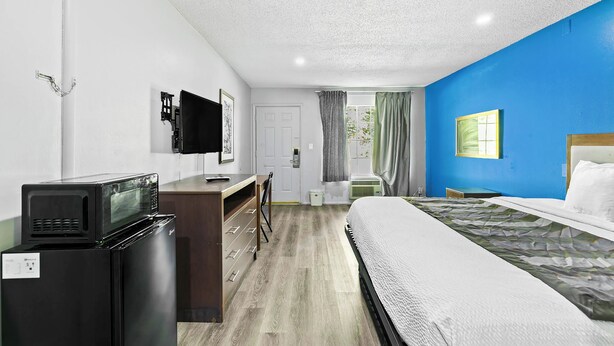 Quarto de hotel com cama, escrivaninha com computador, micro-ondas, televisão e janela com cortinas.
