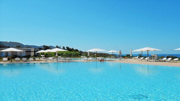 Piscina azul límpida com guarda-sóis brancos e espreguiçadeiras, rodeada por vegetação e vista para o oceano.