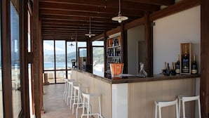 Uma área de bar com teto de madeira, banquetas brancas e vista para o mar através de grandes janelas.