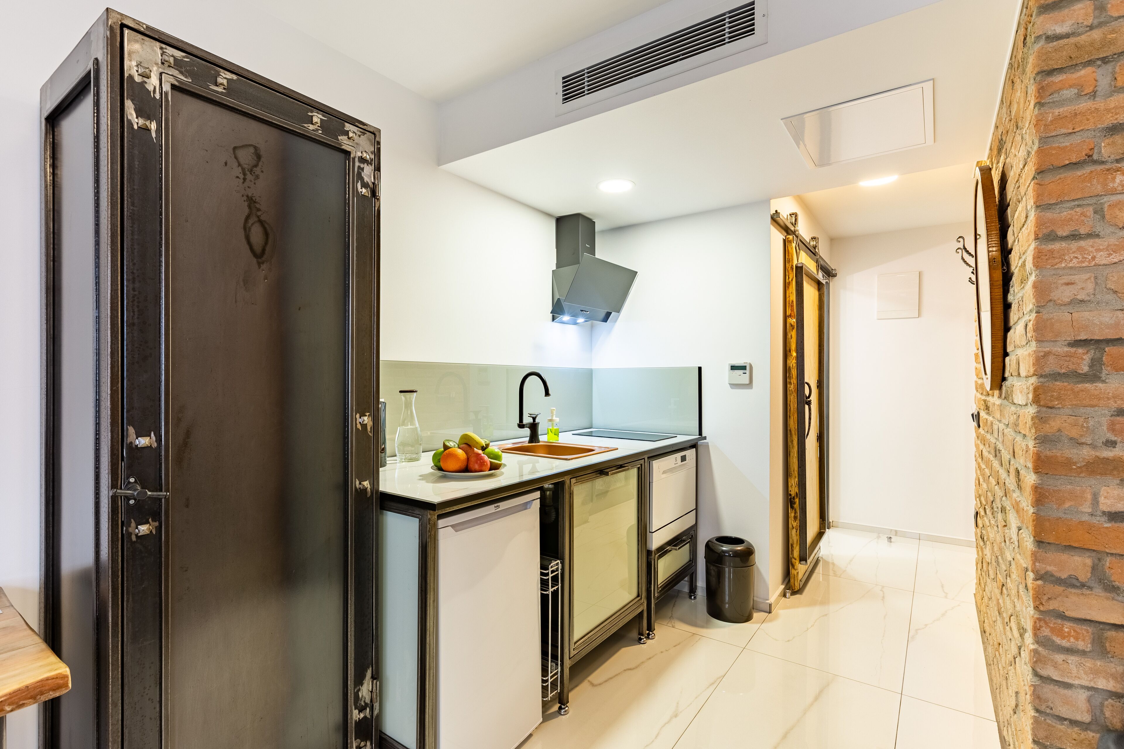 Cozinha compacta com lava-louças, pia e parede de tijolos.