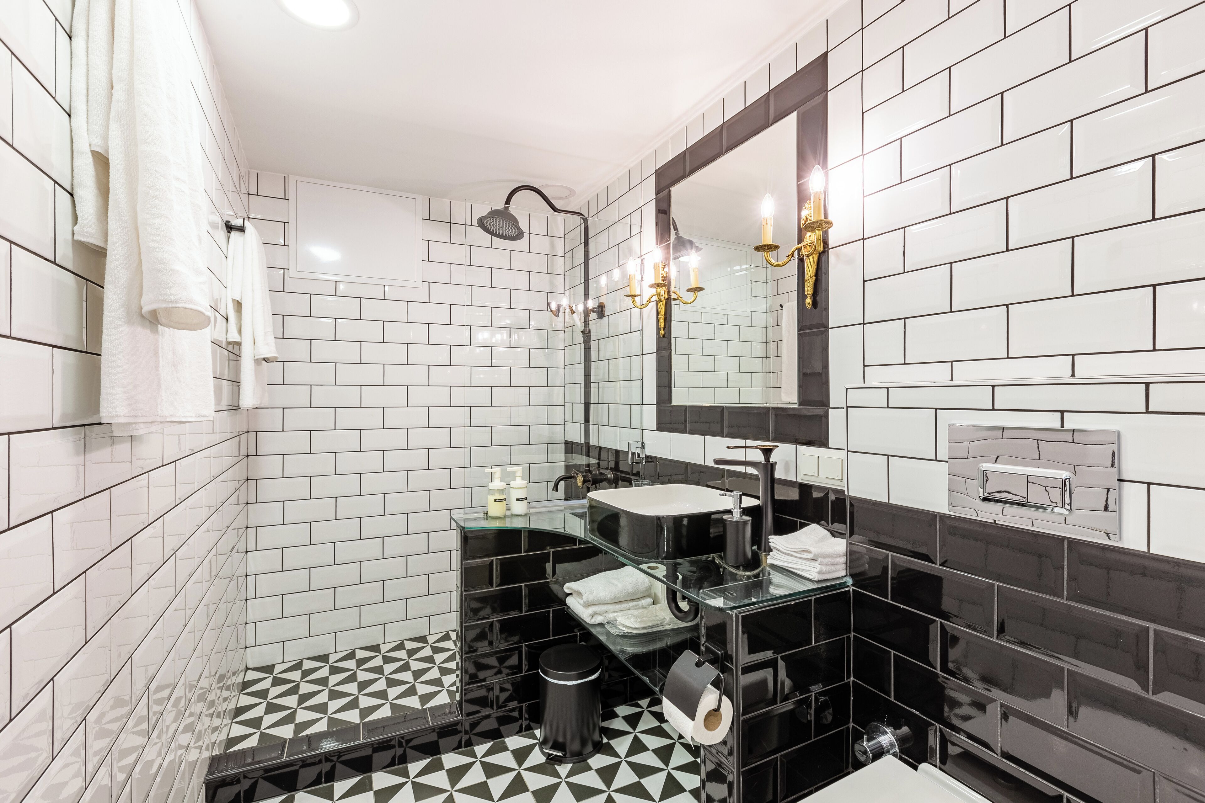 Um banheiro moderno com piso quadriculado em preto e branco, box de vidro, toucador preto com pia de vidro e um espelho grande com arandelas.