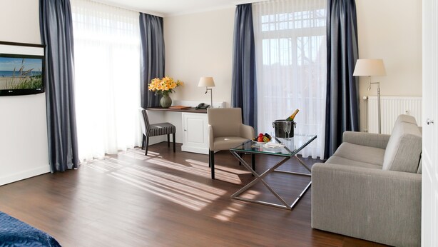 Quarto de hotel moderno com sofá, poltrona e mesa de centro de vidro. Há uma escrivaninha com cadeira e abajur.