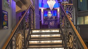 Uma escadaria com grades ornamentadas e degraus iluminados, conduzindo a um espaço com teto iluminado em azul e um candelabro.