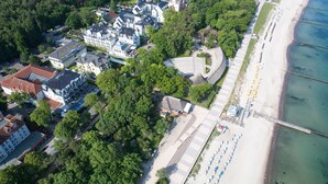 Vista aérea de uma cidade costeira com praia, estação de trem e diversos edifícios, incluindo um hotel com vários andares e um estilo arquitetônico marcante.