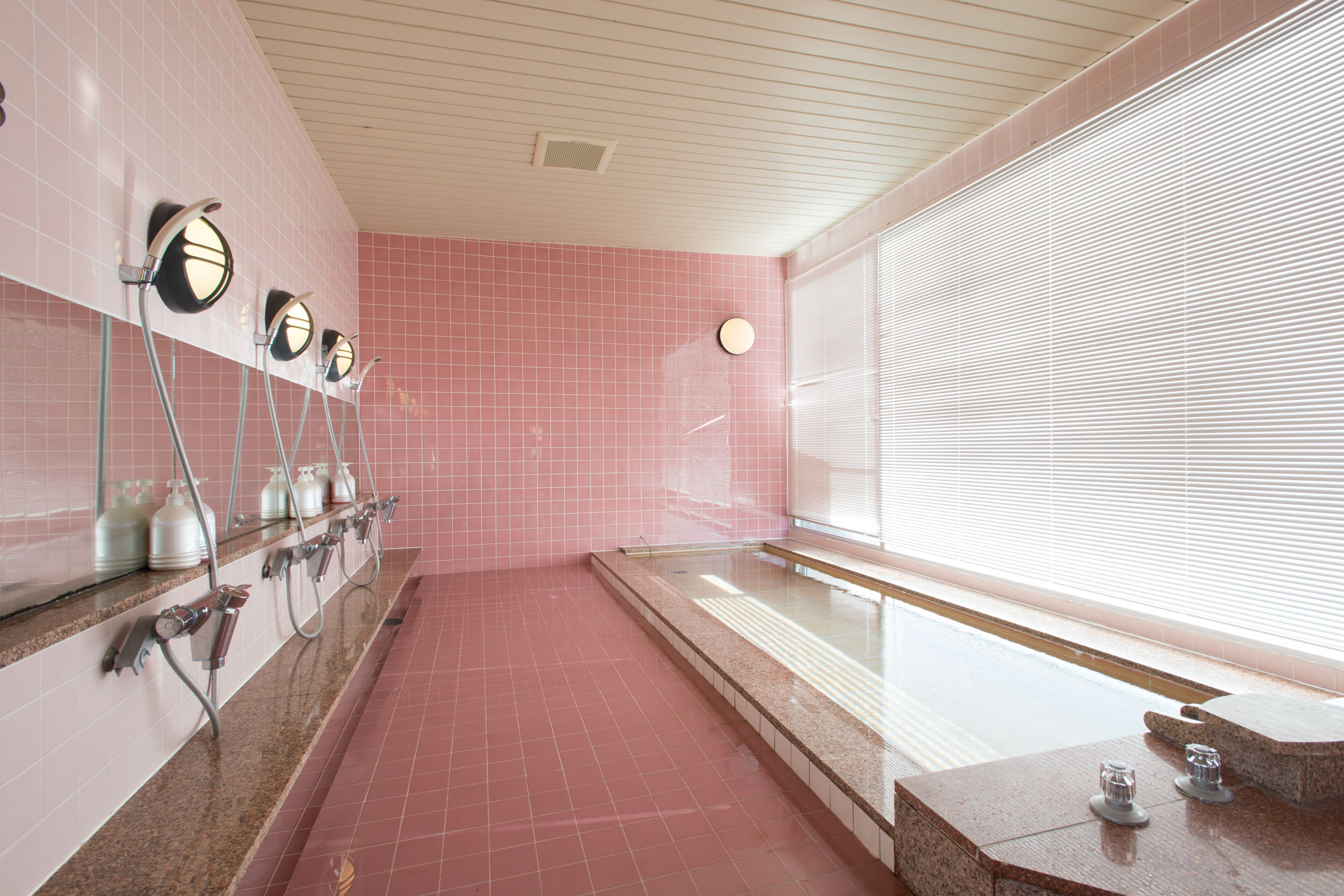 Casa de banho com azulejos cor-de-rosa, cabine de chuveiro em vidro e janela com persianas.