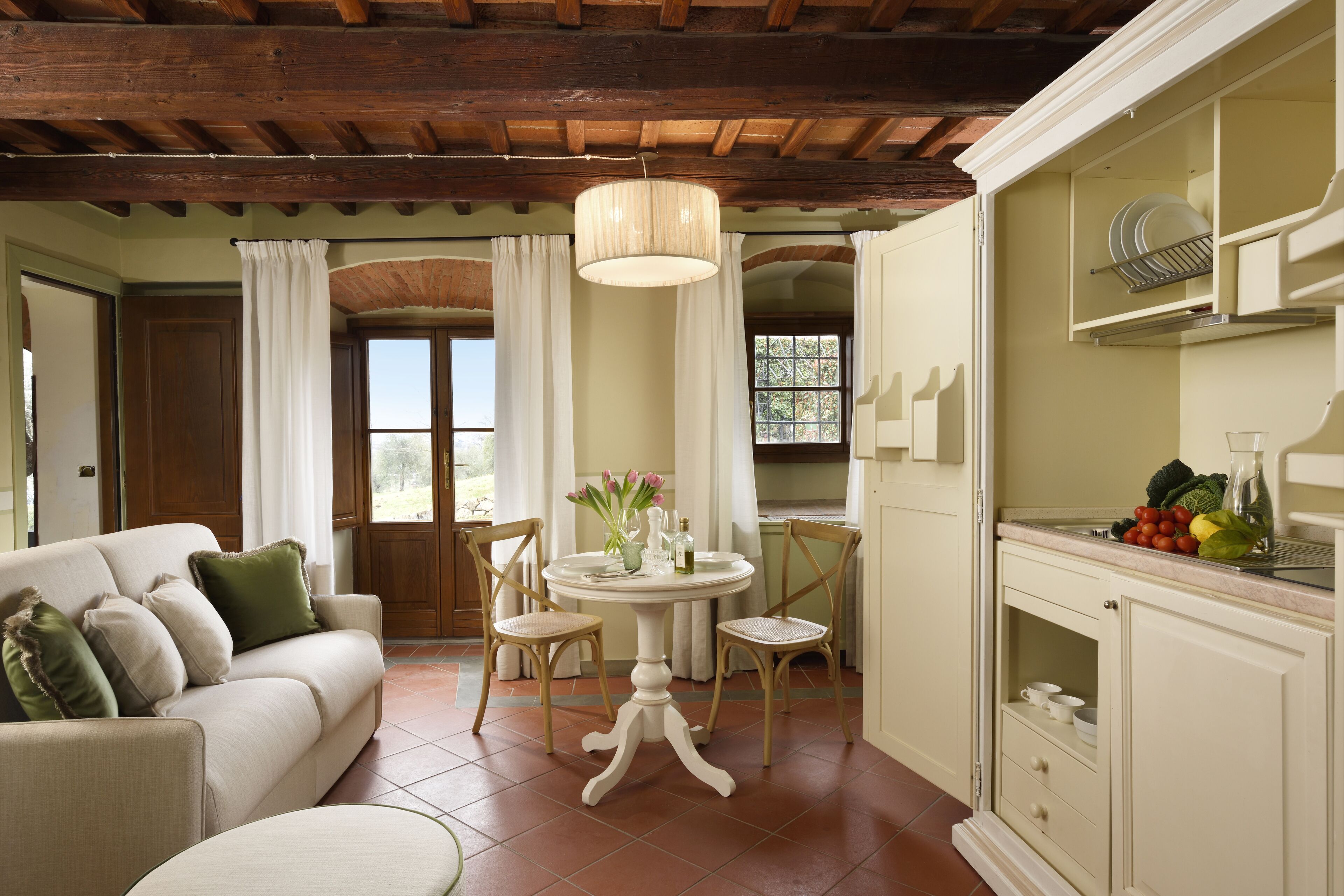 Uma sala de estar acolhedora com um sofá, duas poltronas, uma mesa redonda com um vaso de flores e uma área de cozinha com armários e uma pia.