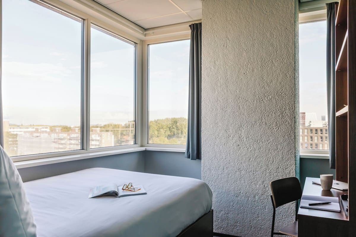 Quarto de hotel com cama, escrivaninha e cadeira. Janelas amplas oferecem vista para edifícios e árvores.