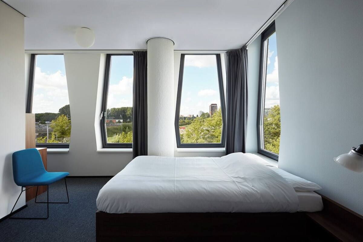 Quarto de hotel com uma cama, uma cadeira, janelas amplas e vista para a cidade.
