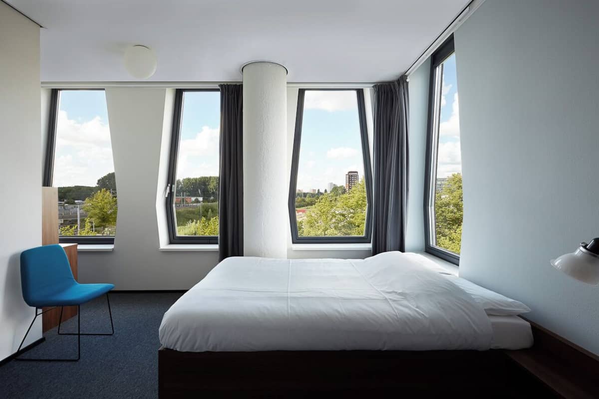 Quarto de hotel com uma cama, uma cadeira, janelas amplas e vista para a cidade.