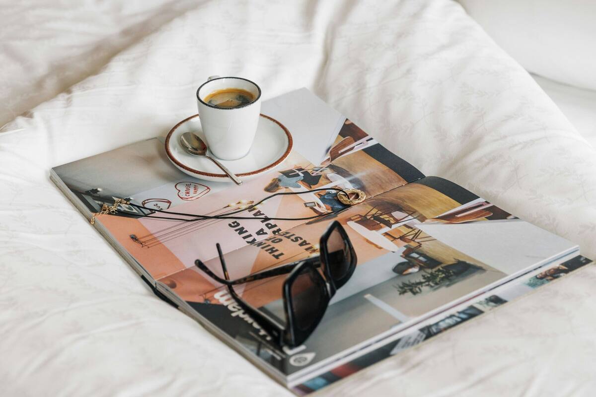 Uma cama com roupa de cama branca, uma xícara de café e uma revista em cima.