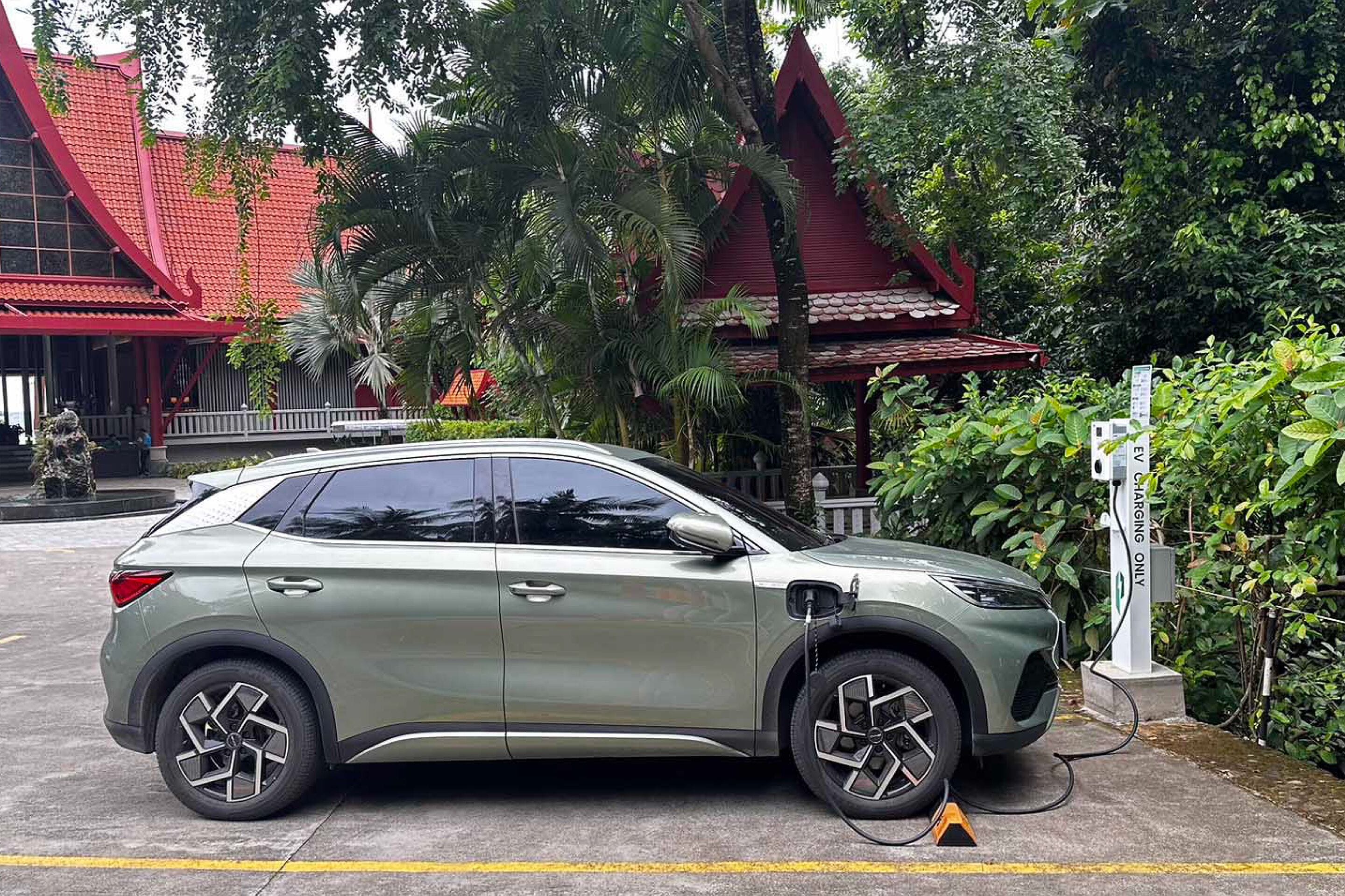 Um SUV verde estacionado em uma estação de carregamento para veículos elétricos, com um edifício de telhado vermelho ao fundo.