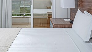 Quarto de hotel com cama, mesa de cabeceira com abajur, porta de vidro deslizante para uma varanda e vista da cidade.