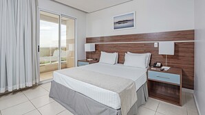 Quarto de hotel com uma cama grande, cabeceira de madeira, mesa de cabeceira com abajur e vista para o exterior através de uma porta de correr.