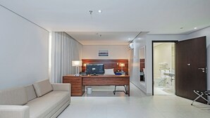 Quarto de hotel moderno com sofá, escrivaninha com computador, cama e banheiro visível por uma porta aberta.