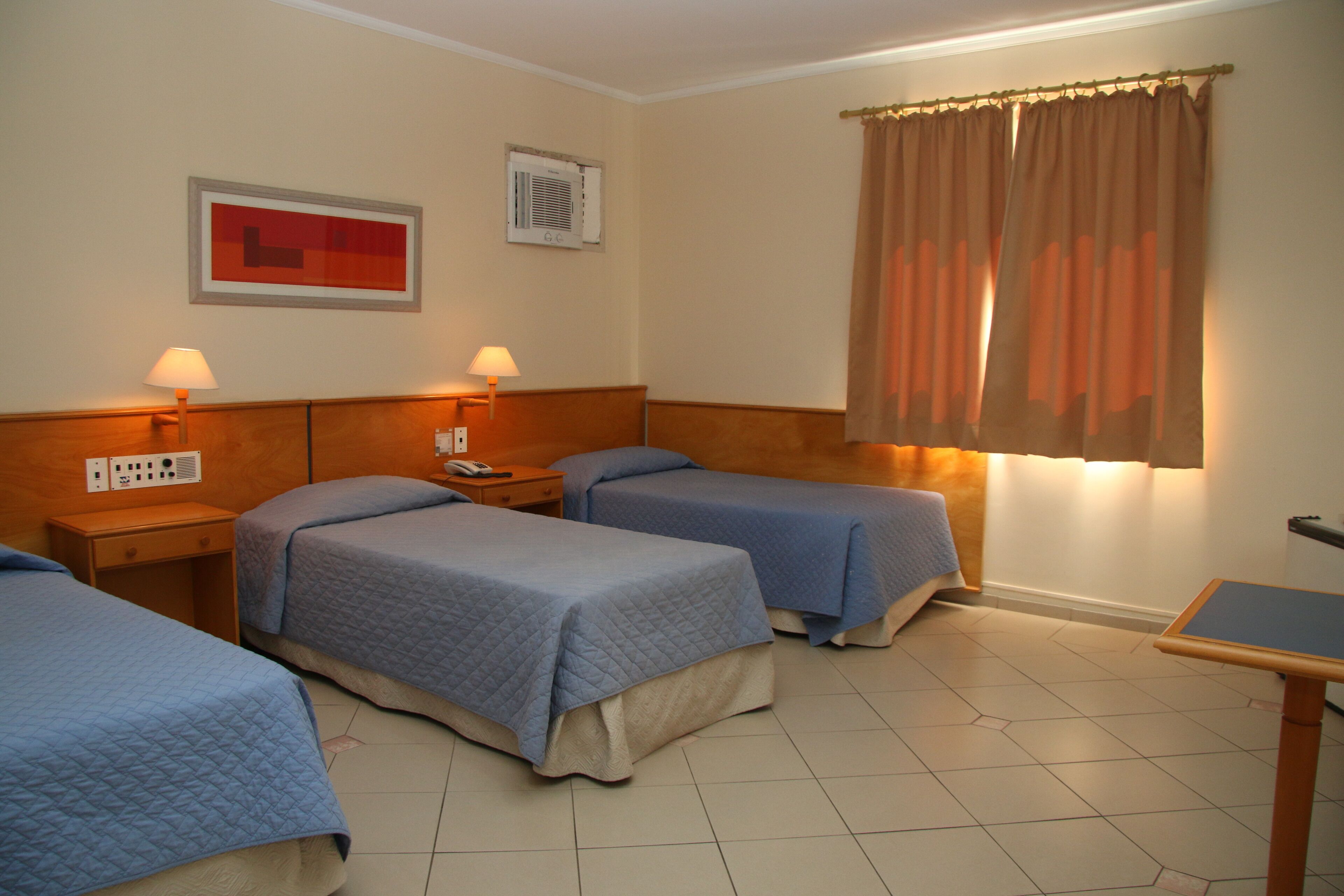Quarto de hotel com duas camas, cabeceira de madeira, uma mesinha e uma janela com cortinas.