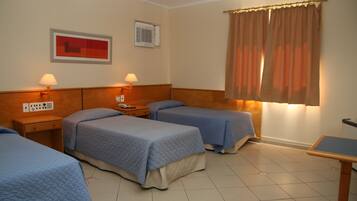 Quarto de hotel com duas camas, cabeceira de madeira, uma mesinha e uma janela com cortinas.