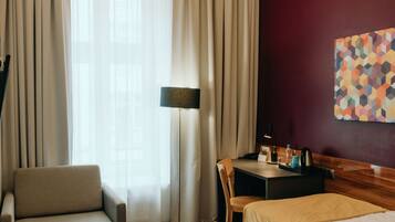 Quarto de hotel com cama, escrivaninha com cadeira, sofá, candeeiro e um quadro na parede.